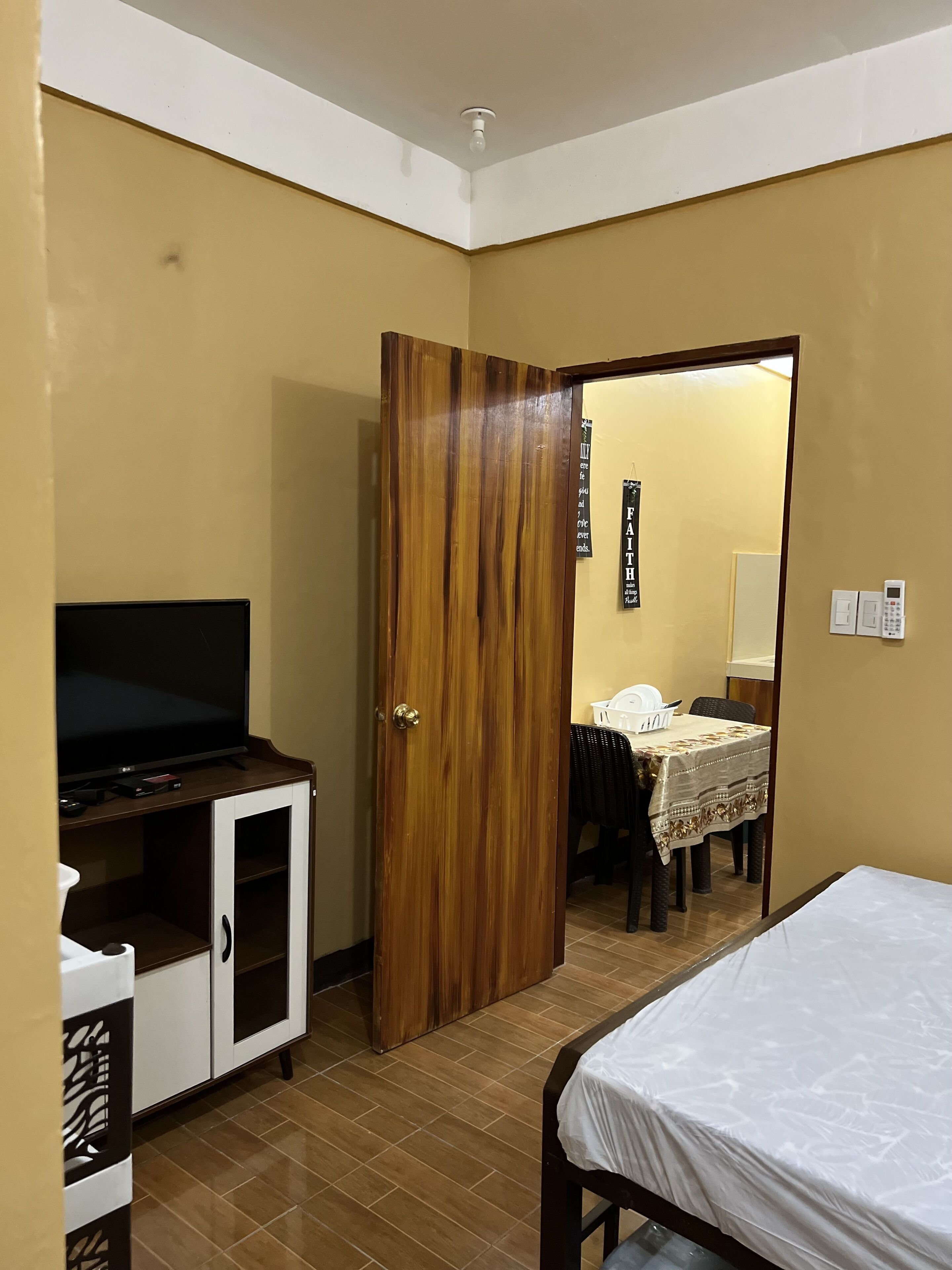 Quarto com guarda-roupa de madeira, televisão sobre um móvel, mesa de jantar com cadeiras e uma cama.