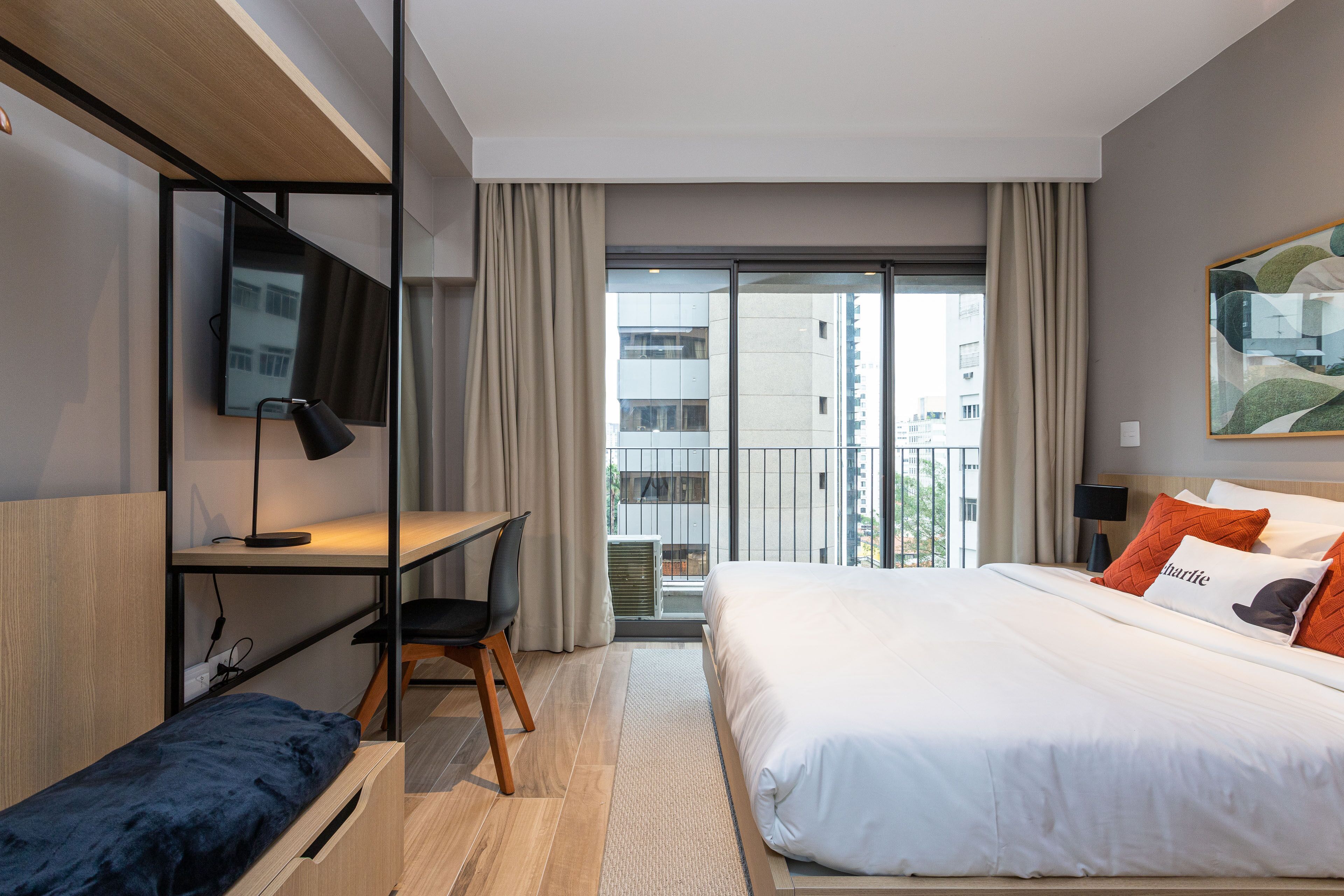 Quarto de hotel moderno com uma cama grande, uma escrivaninha, uma cadeira, uma televisão e uma varanda com vista para a cidade.