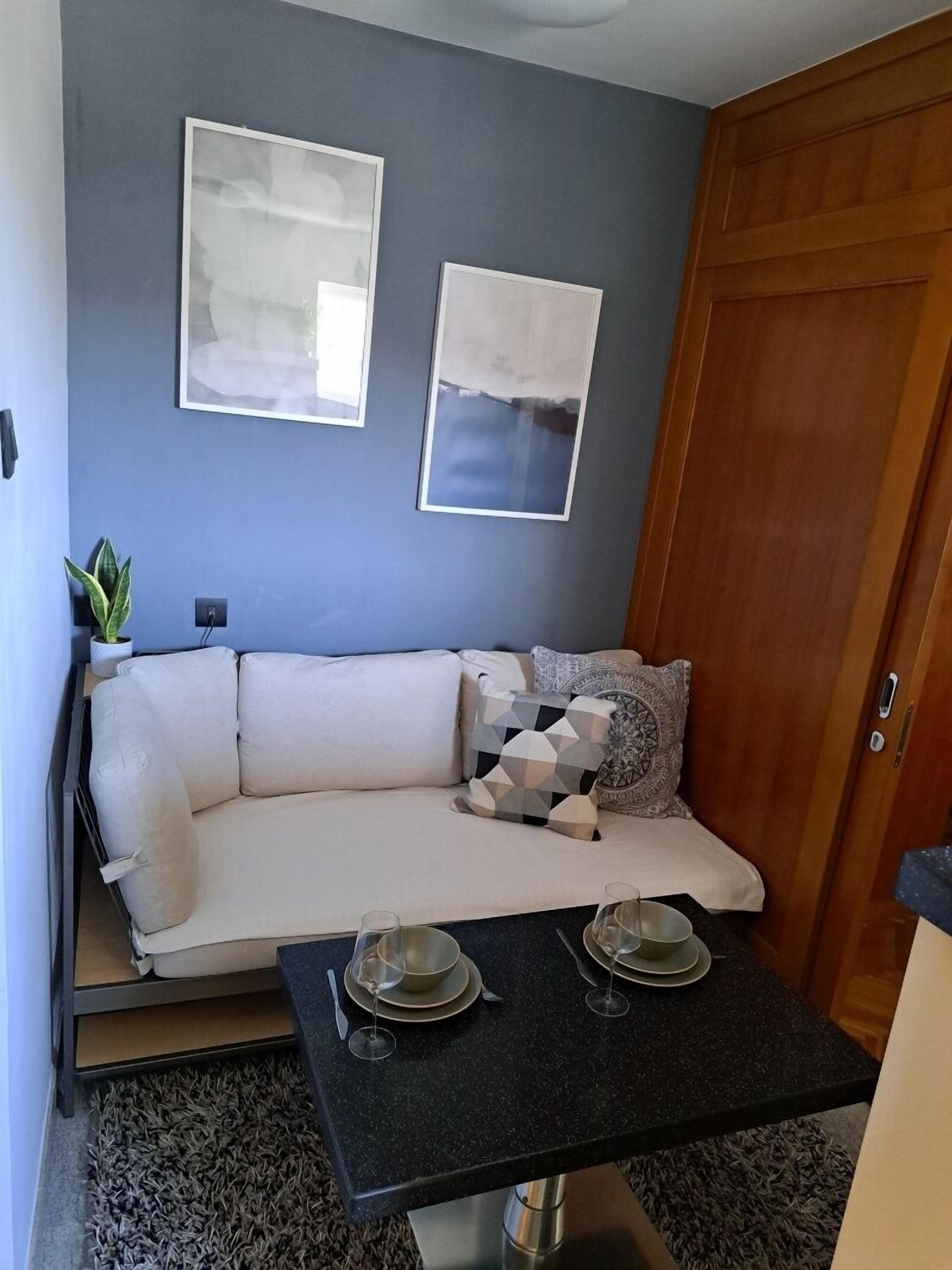 Quarto pequeno e moderno com sofá branco, duas obras de arte abstratas na parede, porta de madeira e mesa de jantar com copos de vinho e pratos.