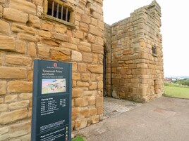 Um edifício de pedra com uma placa de identificação indicando os horários de funcionamento e os preços de entrada para o Mosteiro e Castelo de Tynemouth.