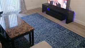 Uma sala de estar com televisão em um suporte preto, uma mesa de jantar, um tapete azul estampado e uma janela com persianas.