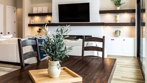 Uma sala de estar moderna com uma mesa de jantar de madeira escura, um vaso branco com plantas verdes e uma TV de tela plana montada sobre uma lareira branca.