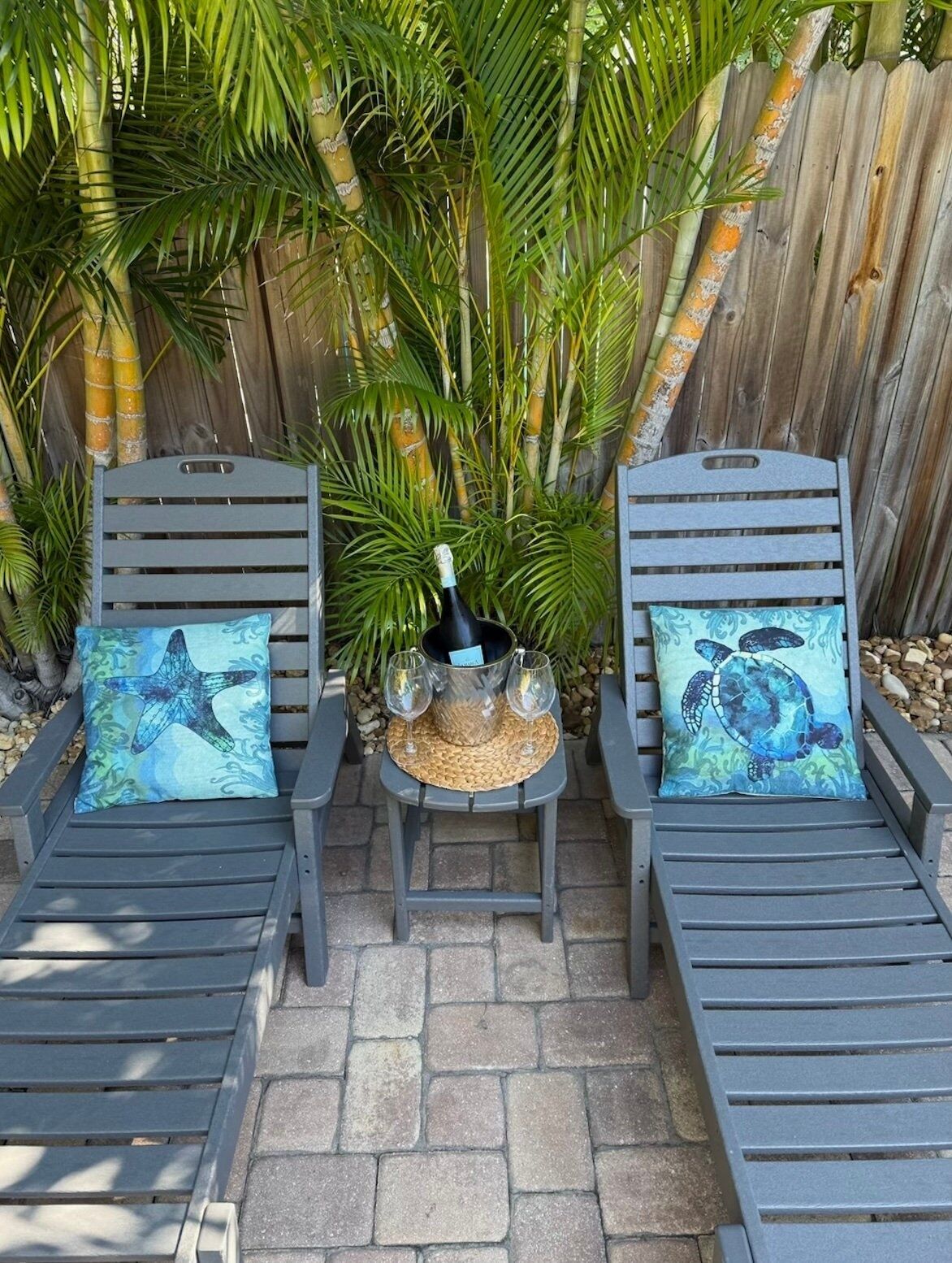 Duas cadeiras de lounge ao ar livre com almofadas combinando, uma mesa pequena com uma garrafa e copos, e um cenário de plantas tropicais e um muro de madeira.