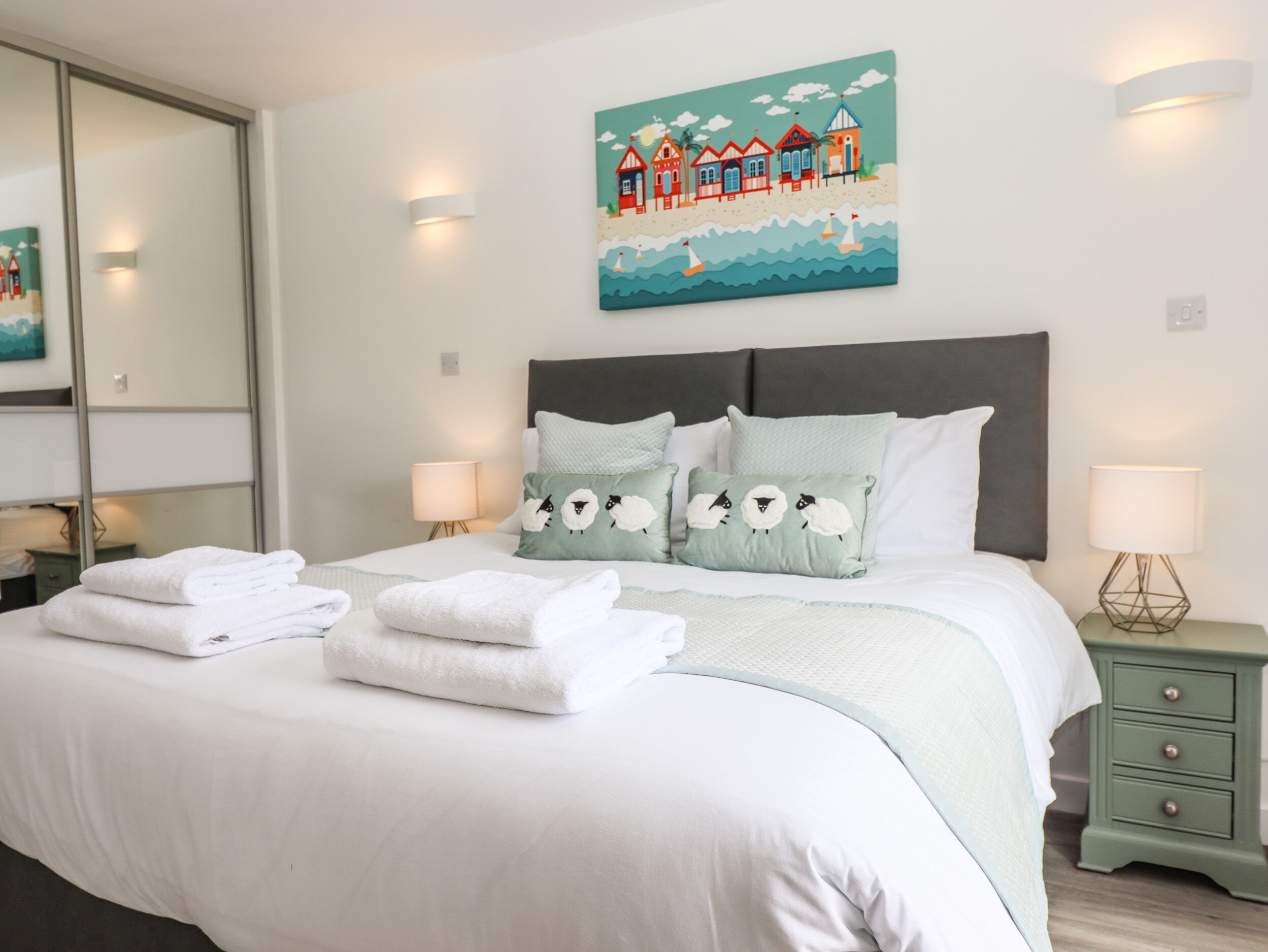 Uma cama bem arrumada com lençóis brancos e um cobertor verde, um quadro de uma paisagem costeira acima dela, e uma mesa de cabeceira com uma luminária e uma gaveta.