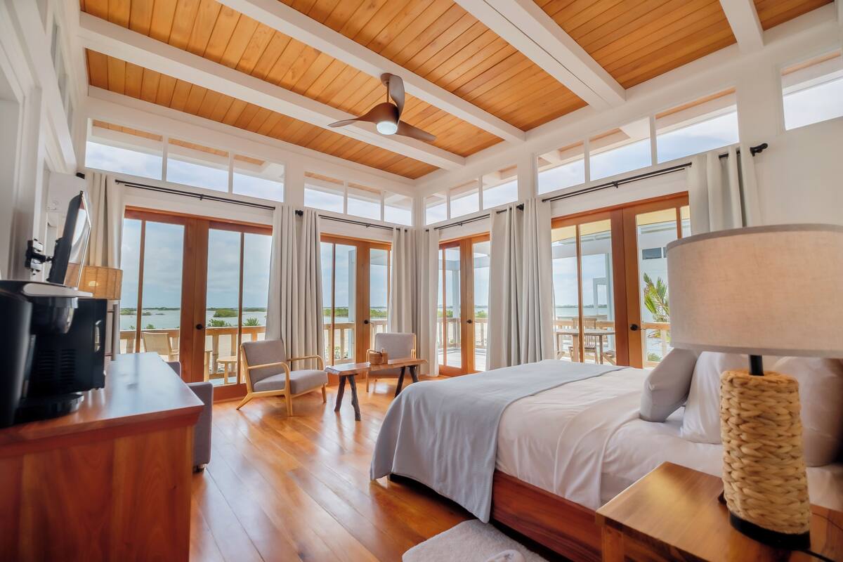 Quarto de hotel com teto de madeira, janelas amplas, uma cama, uma escrivaninha e uma cadeira.