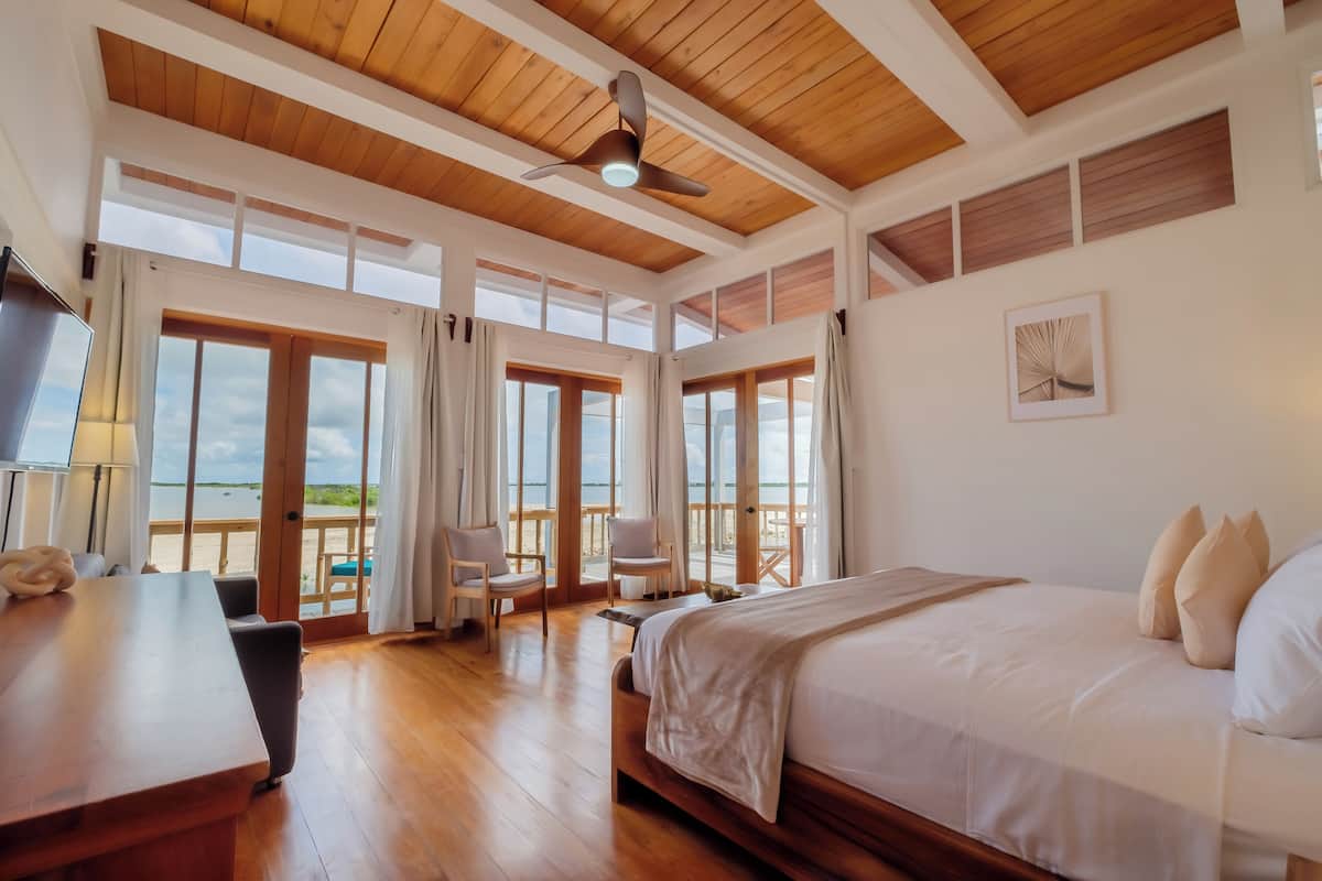 Quarto espaçoso com cama grande, teto de madeira e amplas janelas com vista para a praia.