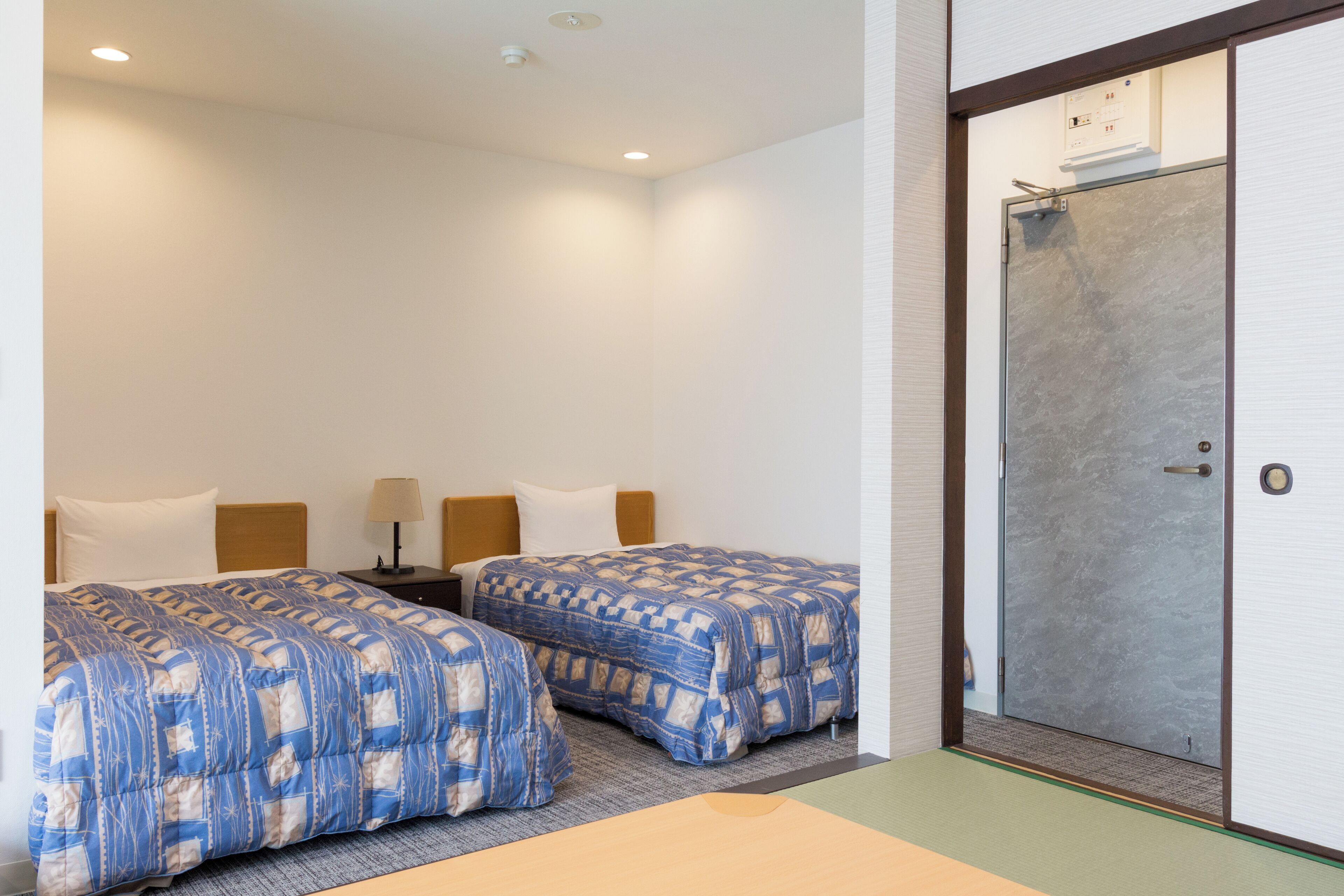 Quarto de hotel com duas camas, área de chuveiro e uma pequena mesa com abajur.
