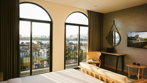Quarto de hotel com uma cama grande, uma escrivaninha com um laptop, uma cadeira e uma mesinha. Há uma janela grande com vista para a cidade.