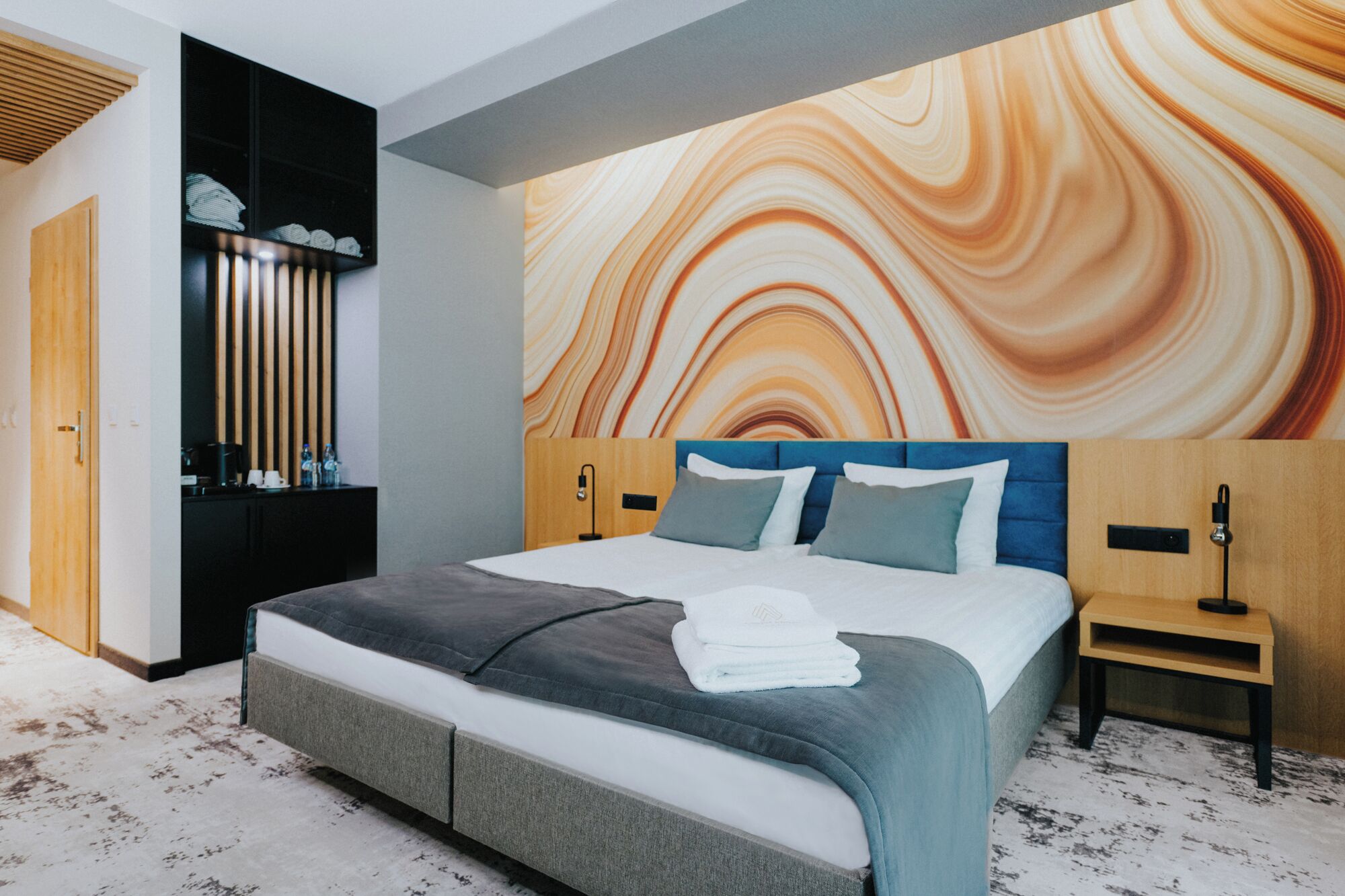Quarto de hotel moderno com uma cama grande, um guarda-roupa de madeira, uma parede com um design abstrato e uma pequena mesa de cabeceira.