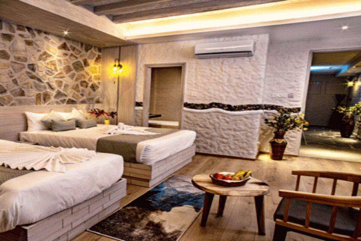 Quarto de hotel com parede de pedra, duas camas, uma mesa pequena com frutas e um banco de madeira.
