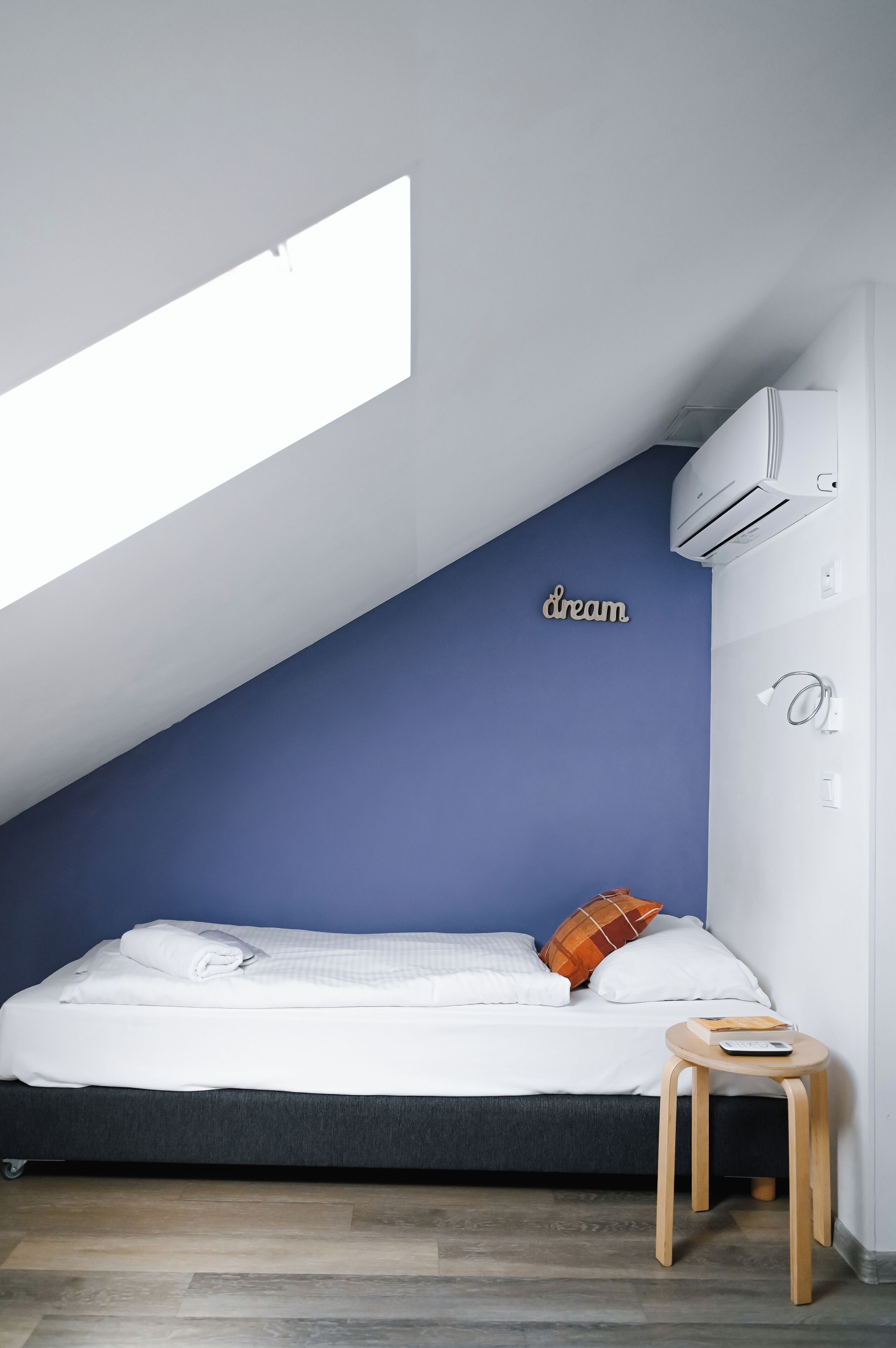 Quarto com teto inclinado, uma cama com roupa de cama branca, um banquinho de madeira pequeno e um aparelho de ar condicionado.