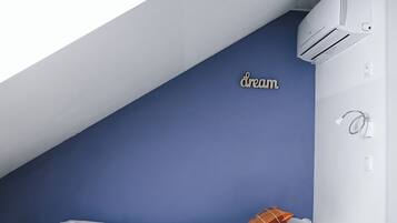 Quarto com teto inclinado, uma cama com roupa de cama branca, um banquinho de madeira pequeno e um aparelho de ar condicionado.