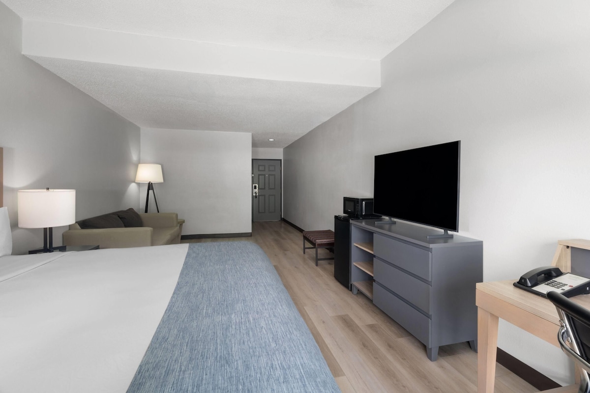 Quarto de hotel com cama, escrivaninha, televisão, abajur, sofá e porta que dá para outro cômodo.