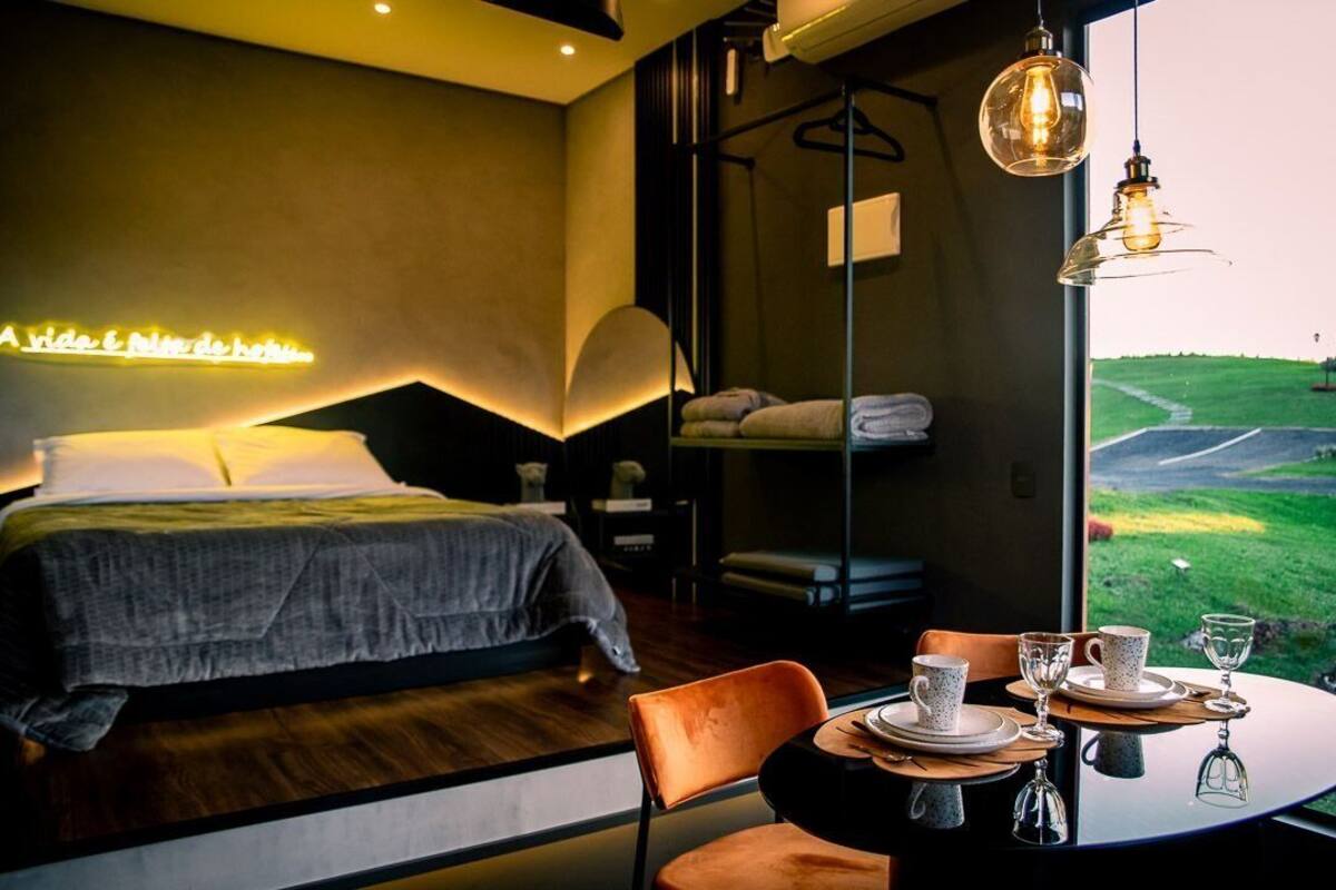 Quarto de hotel moderno com uma cama, uma mesa de jantar com cadeiras e vista para uma paisagem verde através de grandes janelas.