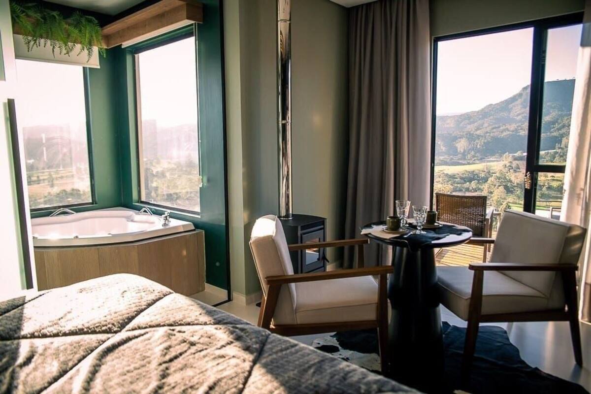 Quarto com cama grande, banheira, área de jantar com cadeiras e vista para paisagem montanhosa.