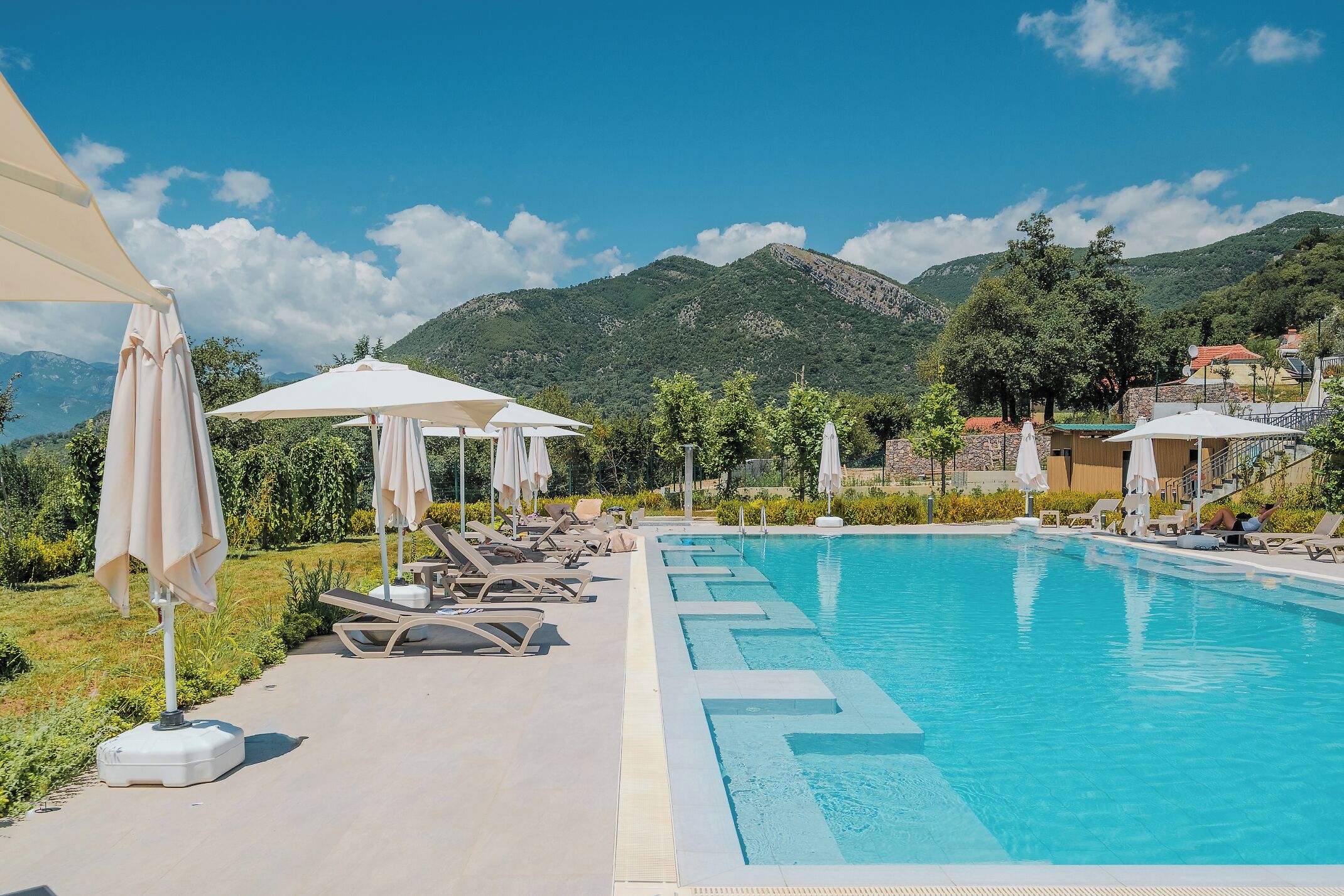 Piscina com espreguiçadeiras e guarda-sóis, cercada por vegetação e montanhas.