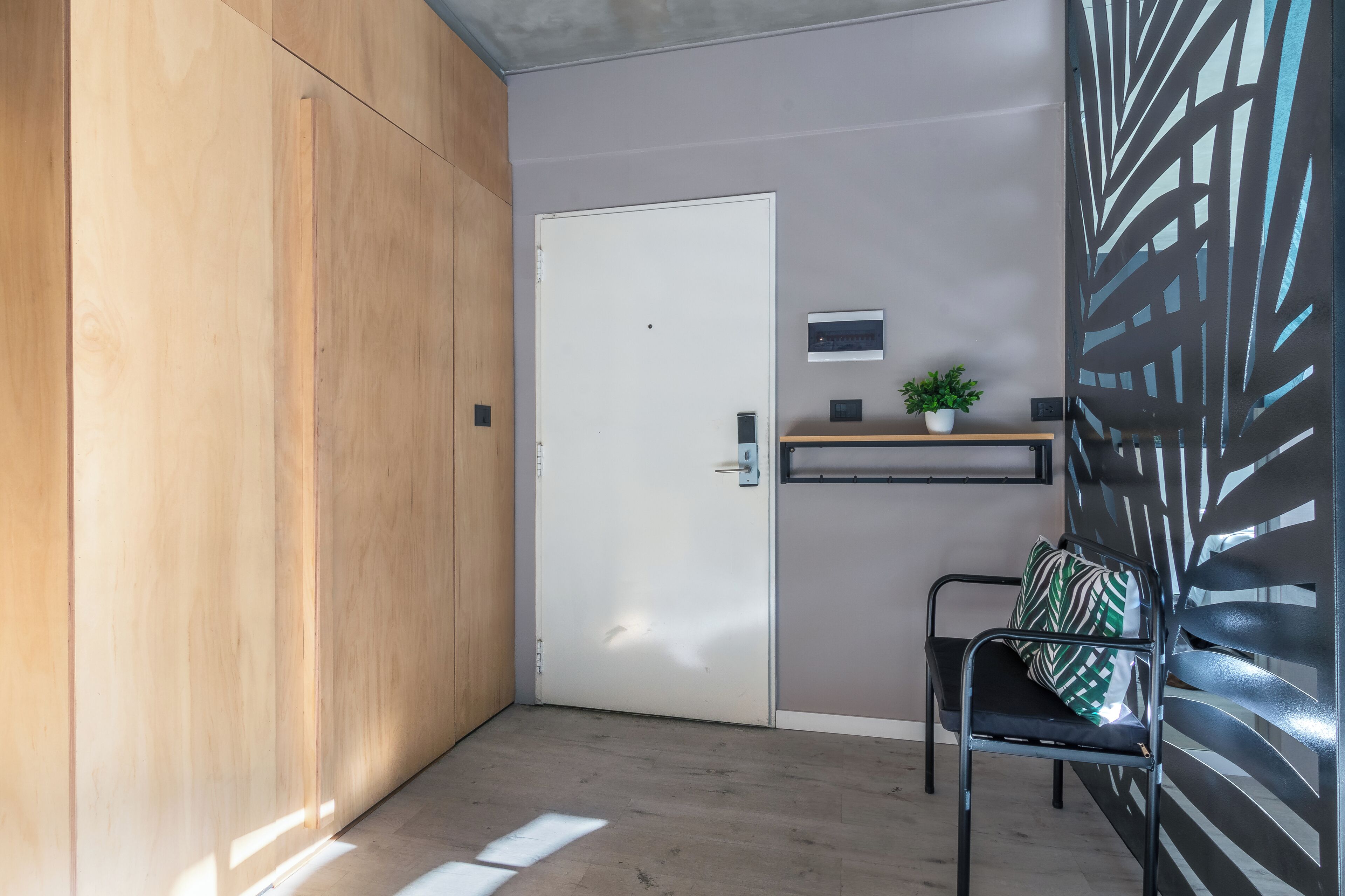 Quarto com guarda-roupa de madeira, porta branca, cadeira preta com almofada estampada, uma pequena planta numa prateleira e parede com design geométrico.