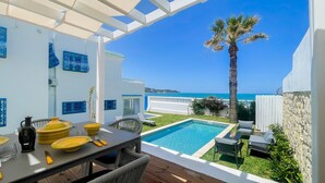 Área da piscina com vista para o mar, mesa de jantar posta com pratos amarelos e uma estrutura de pérgula branca.