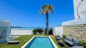 Área da piscina com vista desobstruída para o mar, uma palmeira e móveis de exterior.