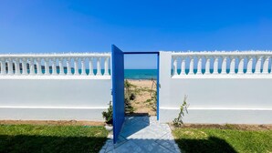 Porta azul aberta com vista para a praia, paredes brancas e céu limpo.
