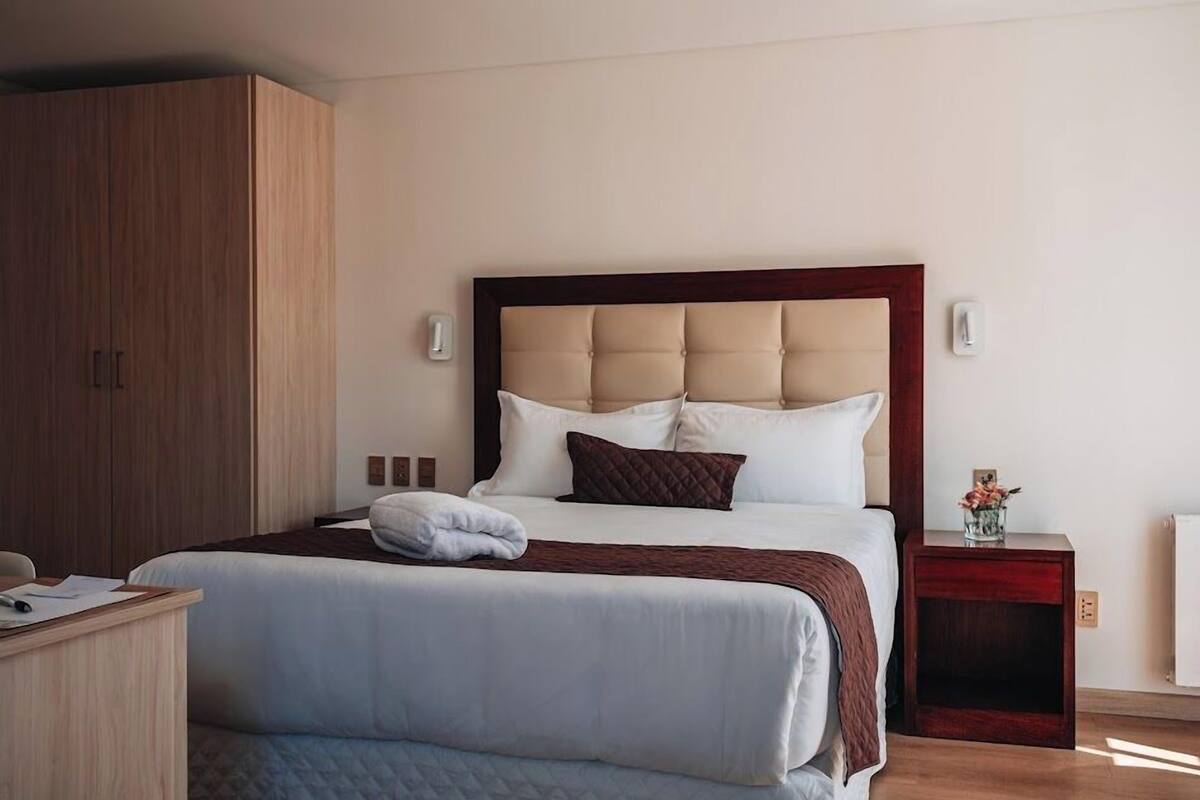 Quarto de hotel com uma cama grande, um guarda-roupa de madeira, uma mesa de cabeceira com um vaso e uma escrivaninha com papéis.