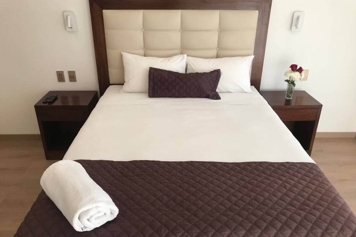 Cama bem arrumada com colcha marrom, travesseiros brancos e toalha branca ao lado. Duas mesas de cabeceira, uma com controle remoto e outra com um vaso de flores.