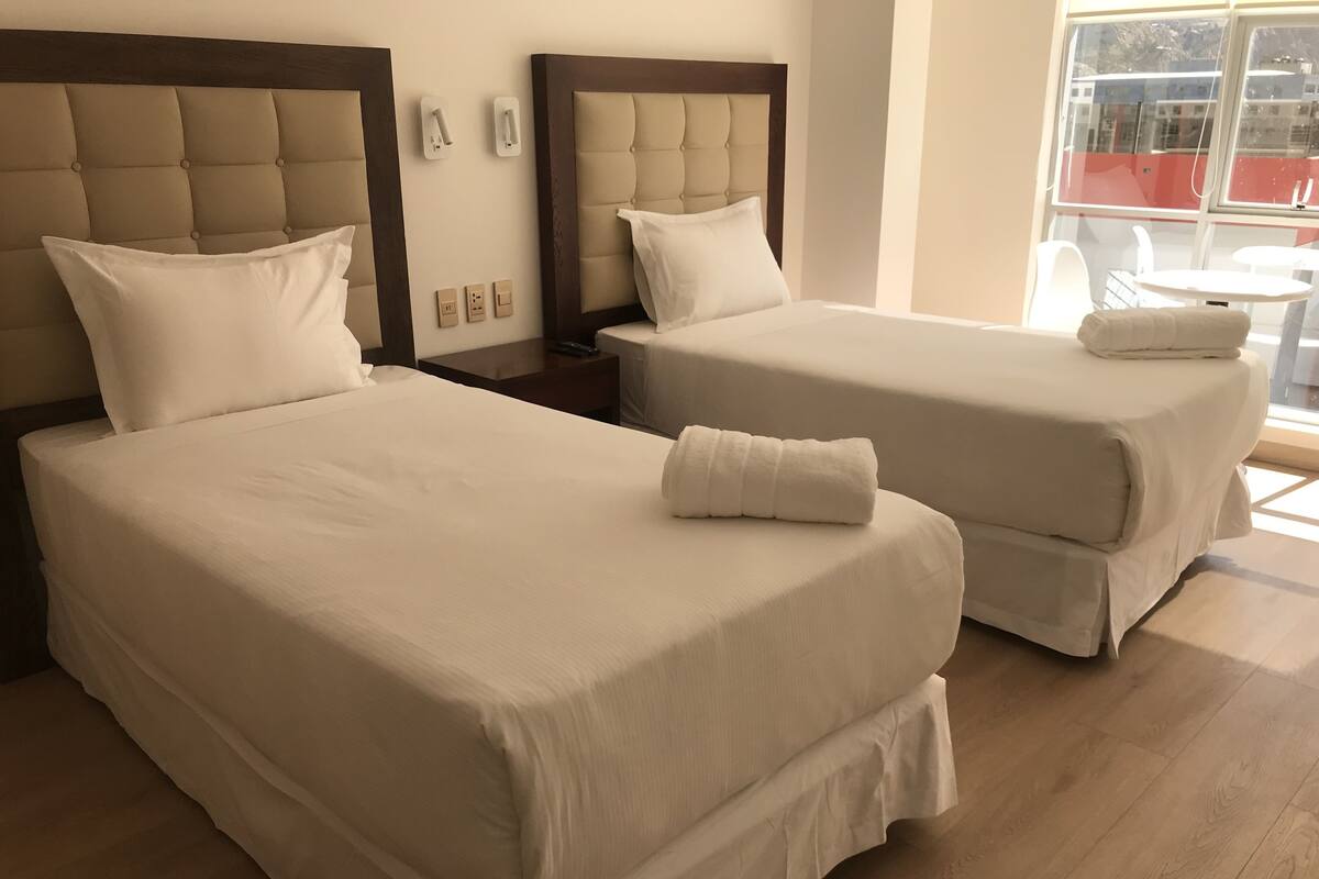 Quarto de hotel com duas camas, cada uma com cabeceira, mesa de cabeceira e janela com vista para o exterior.