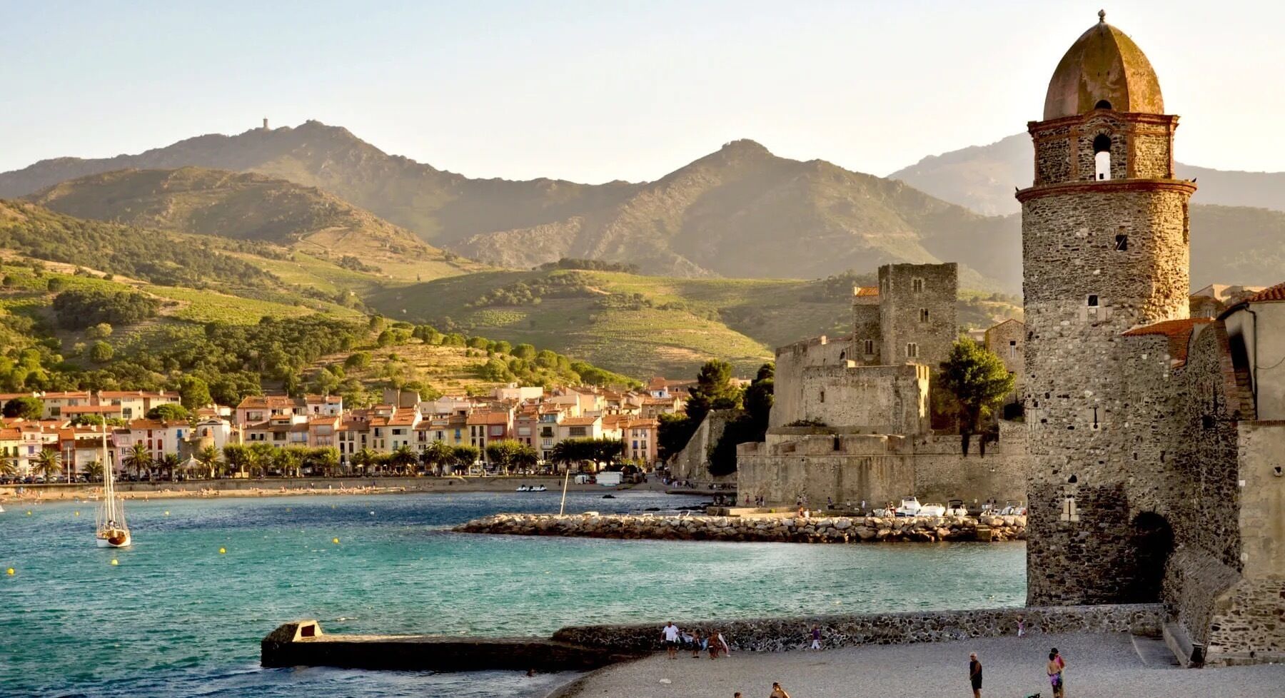 Uma vila costeira com uma torre histórica, uma muralha de pedra e vista clara do mar.