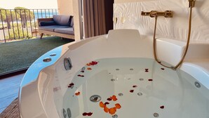 Uma banheira grande e branca com pétalas de rosa, um chuveiro dourado e vista para o mar através de uma varanda.