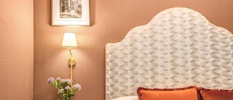 Quarto de hotel com parede texturizada, um quadro emoldurado, mesa de cabeceira com abajur e flores, e uma cama com cabeceira estampada e almofadas.