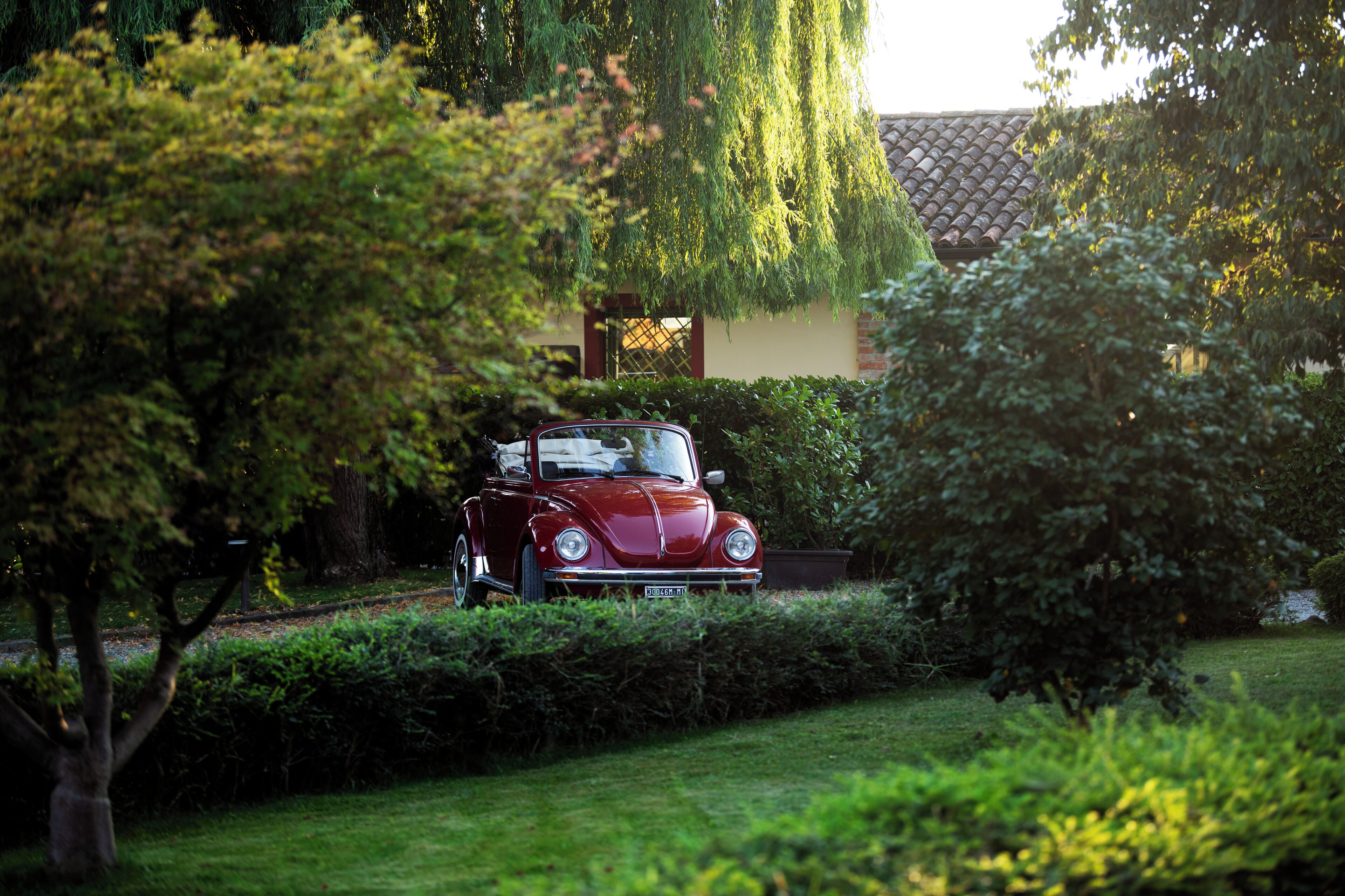 Um carro conversível vermelho clássico estacionado em um jardim verdejante, com uma casa ao fundo.