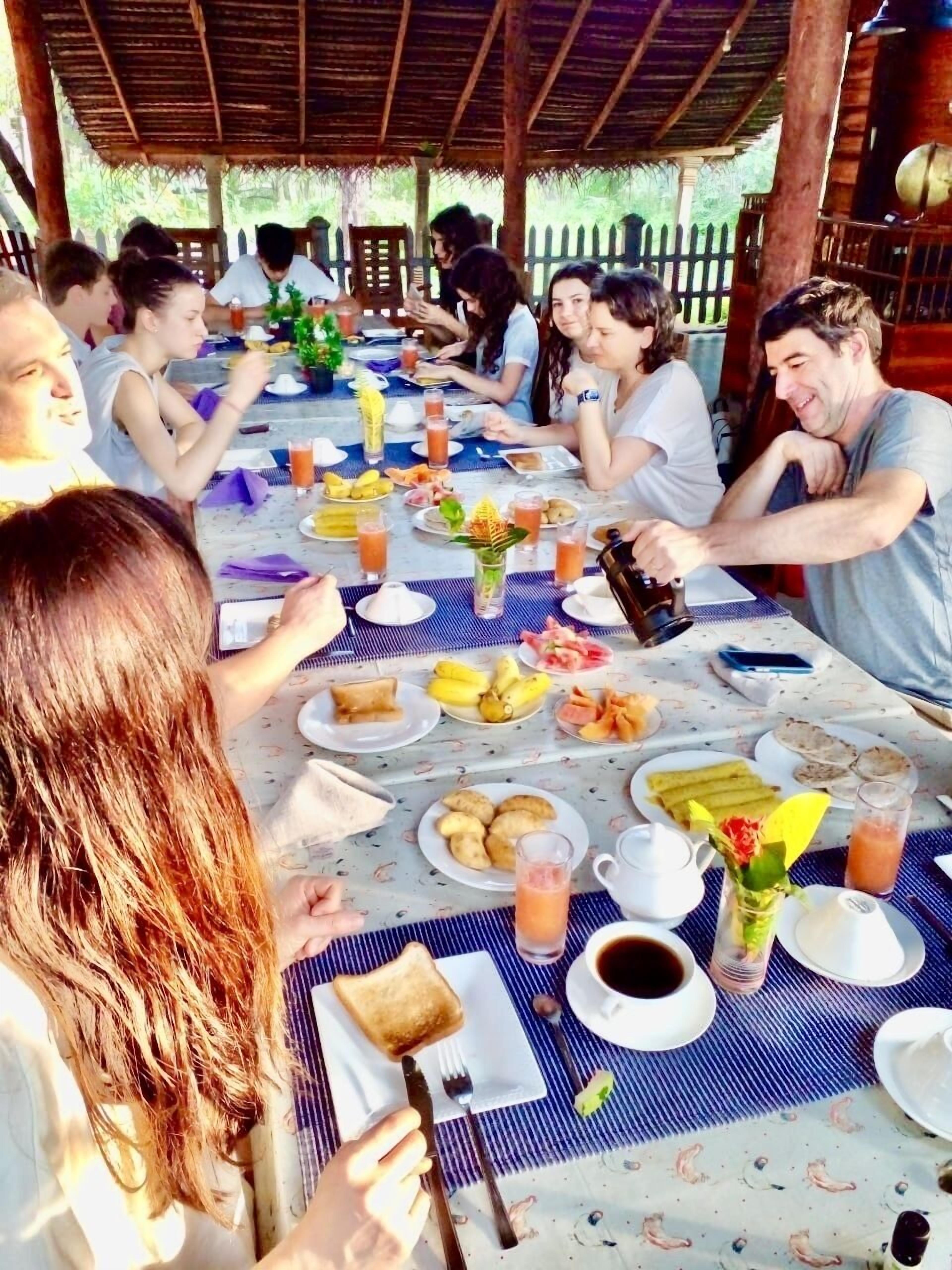 Um grupo de pessoas jantando em uma mesa ao ar livre, com diversos pratos e bebidas.