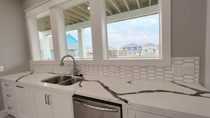 Cozinha com armários brancos, pia de aço inoxidável e lava-louças. Três janelas acima da pia, e um revestimento de azulejos hexagonais.