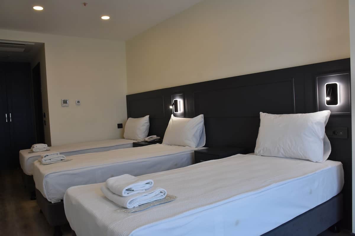 Quarto de hotel com três camas de solteiro, cada uma com colcha e almofadas brancas, e cabeceira escura. Há mesinhas de cabeceira com abajures e um termostato visível na parede.