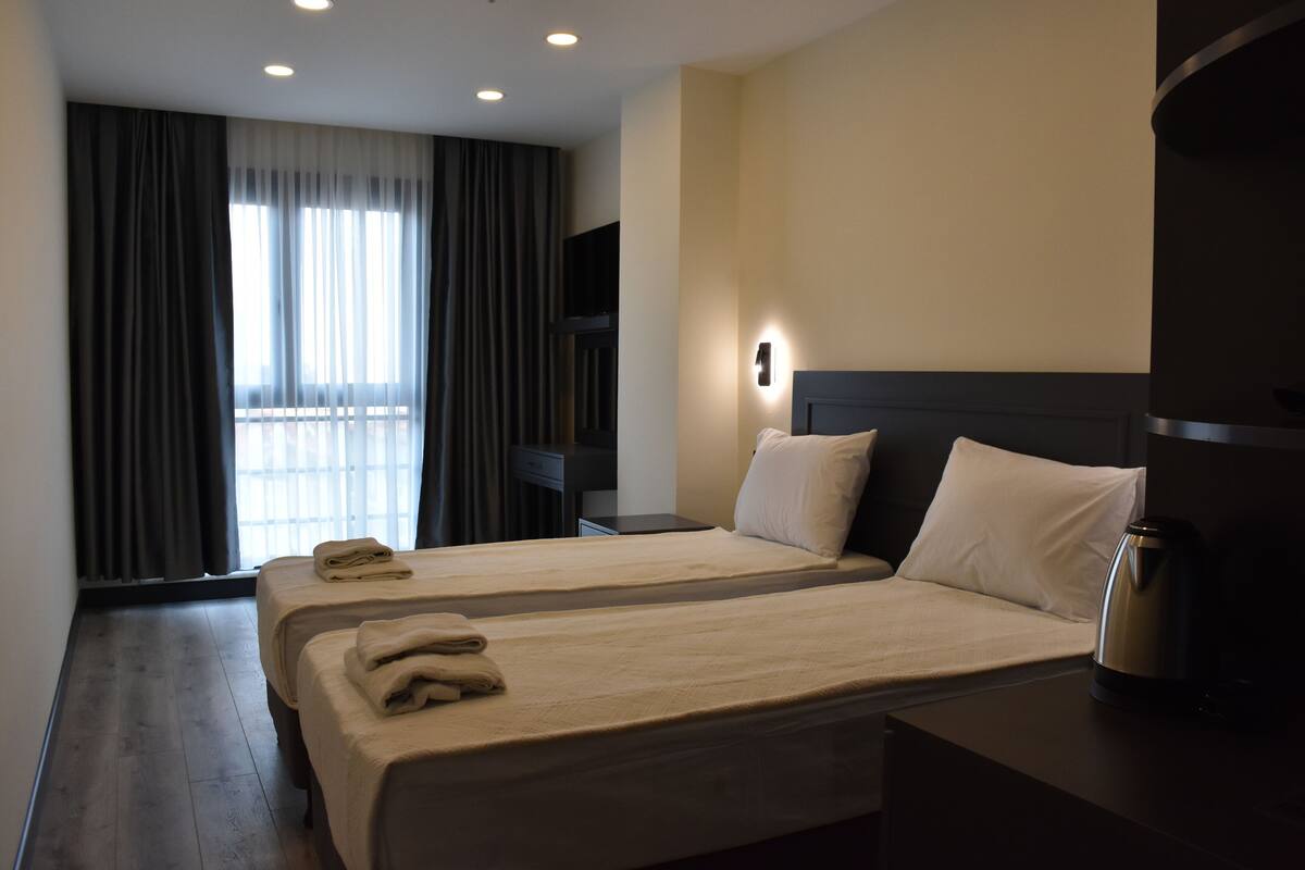Quarto de hotel com duas camas, uma mesinha de cabeceira, uma luminária, um chaleira e uma janela com cortinas.