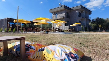 Um edifício com mesas ao ar livre e guarda-sóis, uma área gramada e uma mesa.