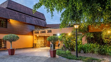 Entrada de um hotel com fachada de pedra, uma placa com a inscrição "PUNE DAK & JNN" e árvores em vasos no primeiro plano.