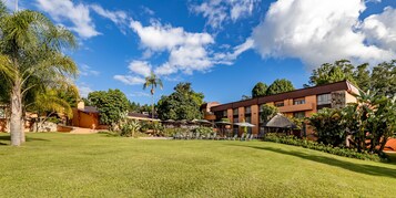 Pátio de hotel com gramado, palmeiras e um edifício com várias varandas e áreas de estar ao ar livre.