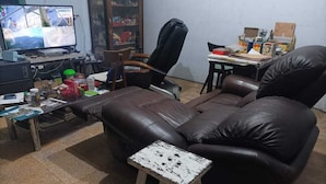 Uma sala de estar com um sofá de couro marrom, um banquinho de madeira, uma televisão exibindo um jogo de vídeo e diversos objetos espalhados pelo chão e pela mesa de centro.