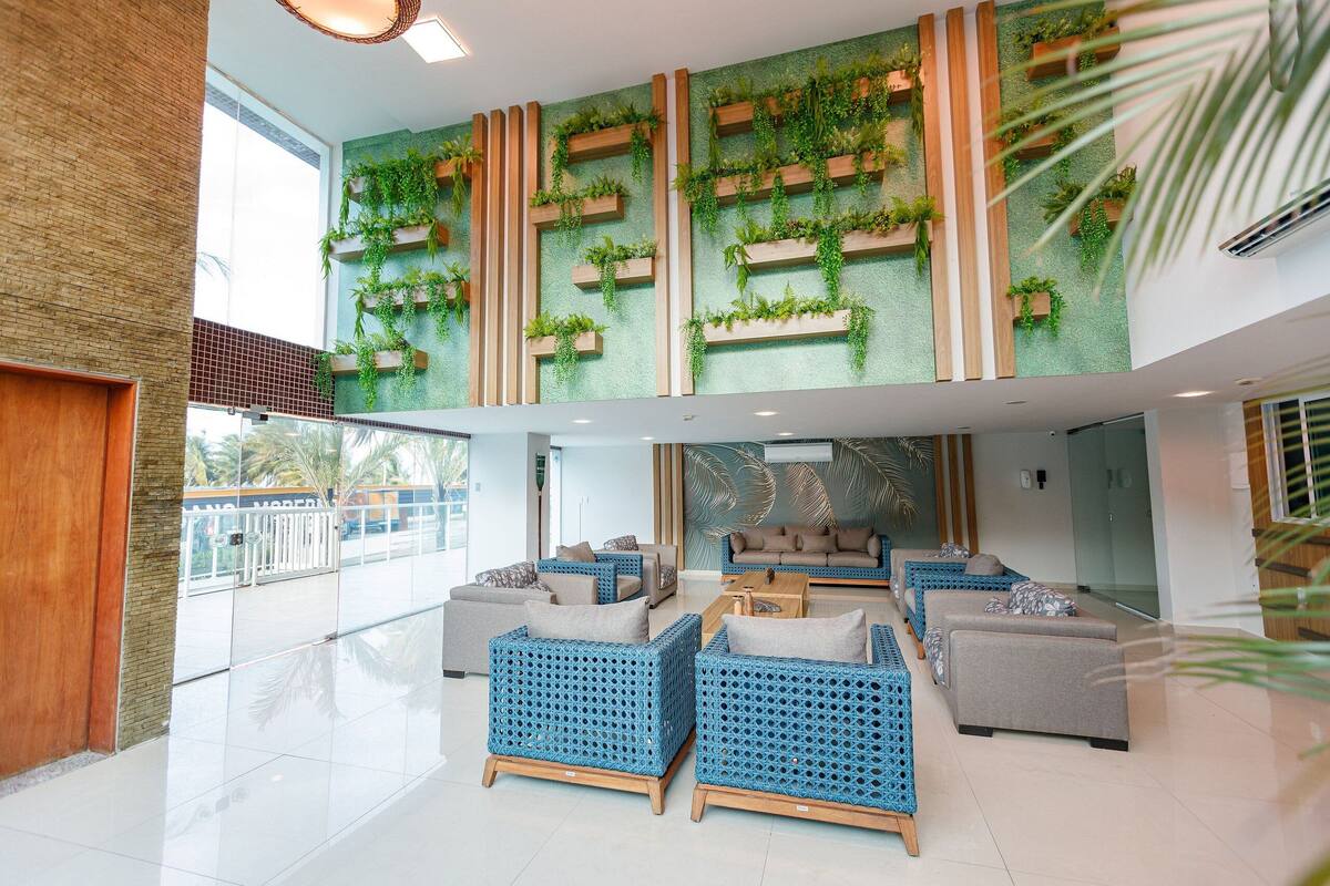 Um lobby amplo com teto alto, área de estar com sofás azul e cinza, e parede verde com jardins verticais.