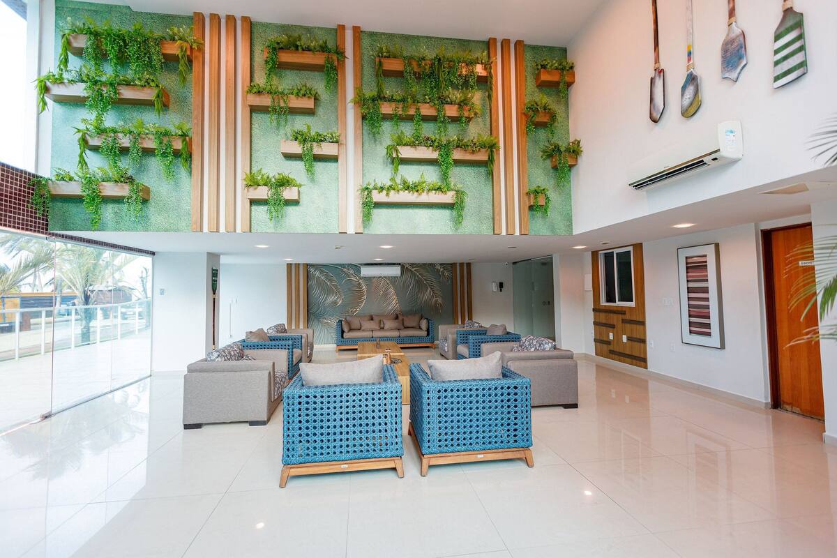 Um hall amplo com teto alto, apresentando uma parede verde com plantas verticais, uma área de estar com sofás de vime azul e piso polido.