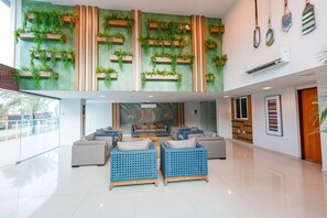 Um hall amplo com teto alto, apresentando uma parede verde com plantas verticais, uma área de estar com sofás de vime azul e piso polido.