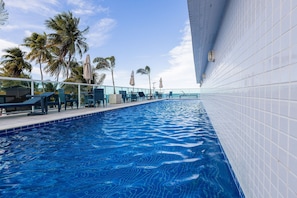 Uma piscina com água azul cristalina, cercada por espreguiçadeiras e guarda-sóis, com palmeiras ao fundo.
