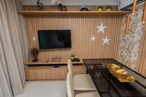 Uma sala de estar moderna com parede de painéis de madeira, televisão de tela plana, mesa de jantar de vidro com cadeiras e elementos decorativos como enfeites em formato de estrela e um tabuleiro dourado.