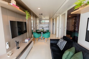 Uma sala de estar moderna com televisão de tela plana, um sofá preto, cadeiras verdes e um espaço de jantar com mesa e cadeiras.
