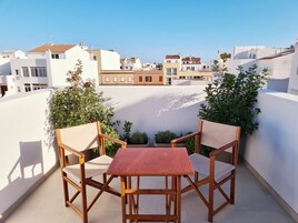 Terraço no telhado com mesa e cadeiras de madeira, rodeado por paredes brancas e vegetação.