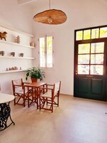 Quarto com mesa de jantar, cadeiras, luminária pendente, planta em vaso e porta com vista para o exterior.