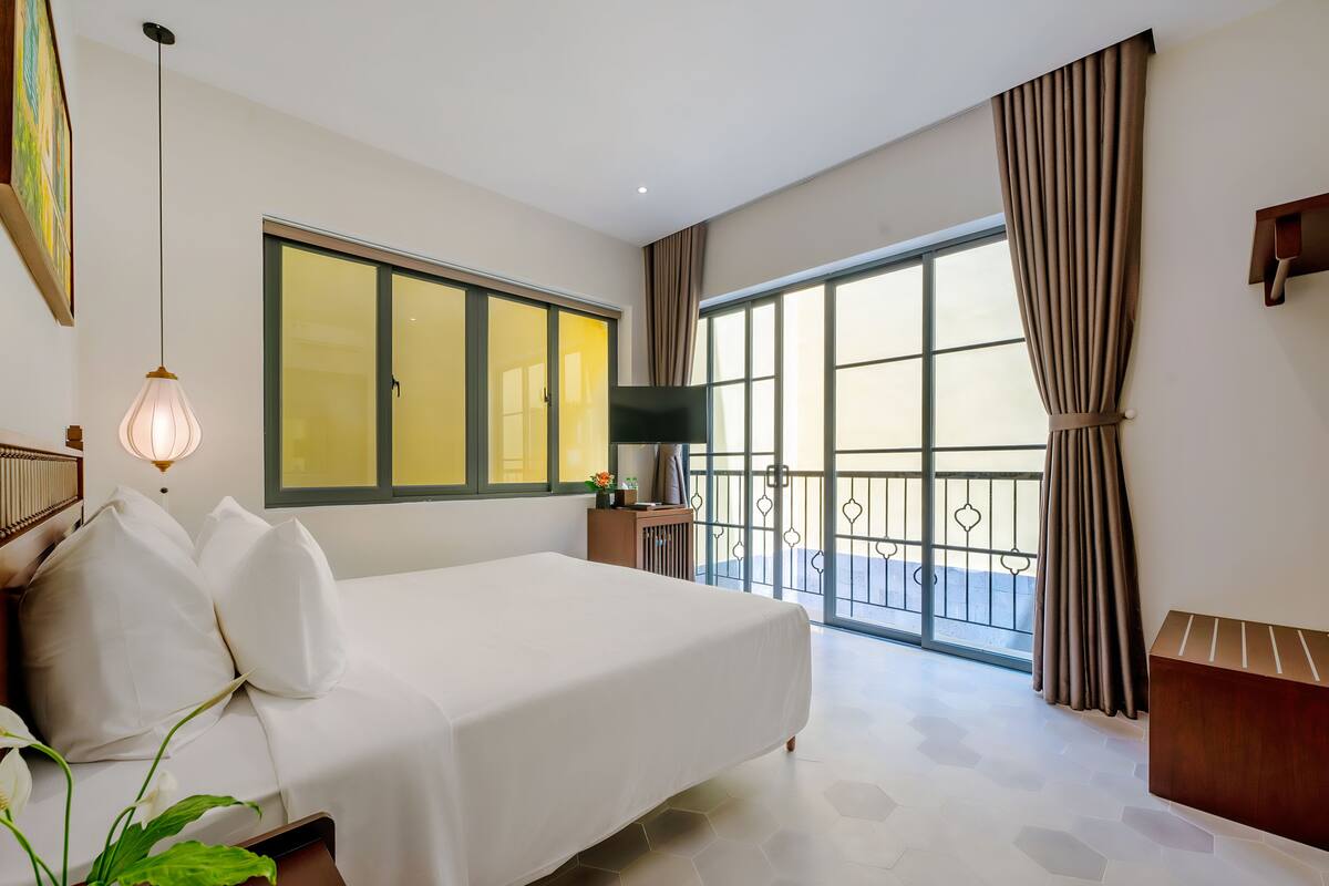 Quarto de hotel com uma cama grande, televisão sobre um suporte de madeira, varanda com vista e uma janela ampla com cortinas.