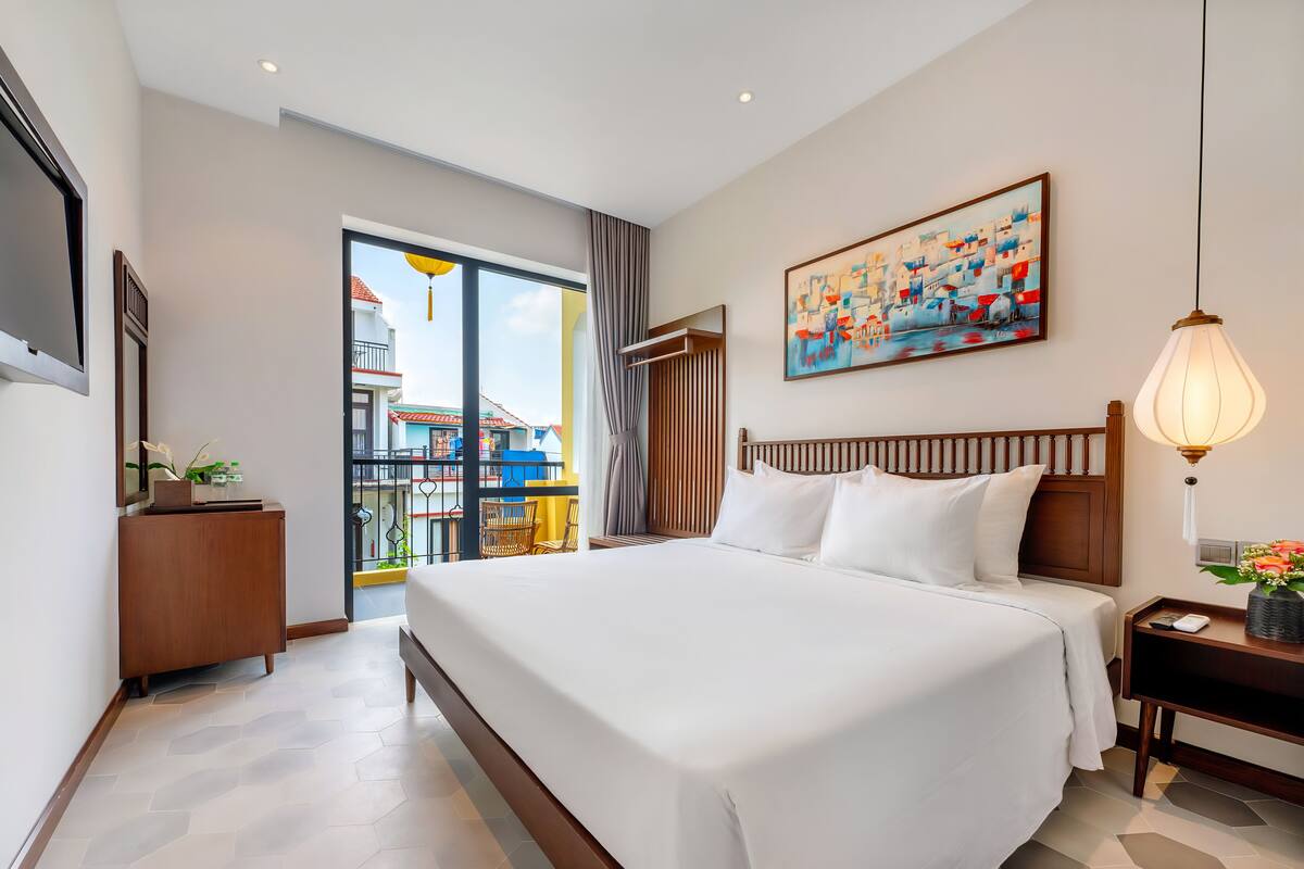 Quarto de hotel com uma cama grande, cabeceira de madeira, mesa de cabeceira com abajur, pintura na parede e vista para a varanda.