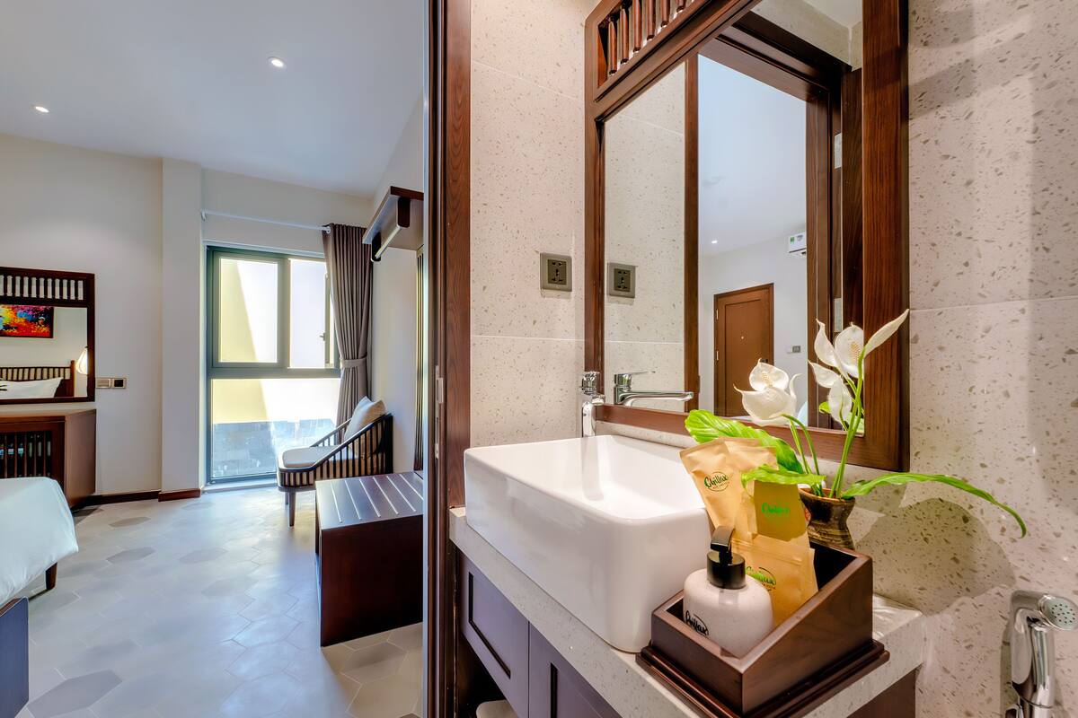 Quarto de hotel com uma pia grande e branca, um espelho e uma orquídea em vaso. Há uma cama e uma cadeira no quarto.
